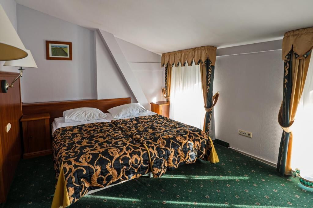 Quarto de hotel com cama, mesa de cabeceira, abajur, quadro na parede e cortinas.