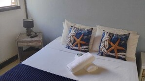Cama com dois travesseiros com estampas de estrelas-do-mar, uma mesinha de cabeceira com abajur e uma toalha enrolada sobre a cama.