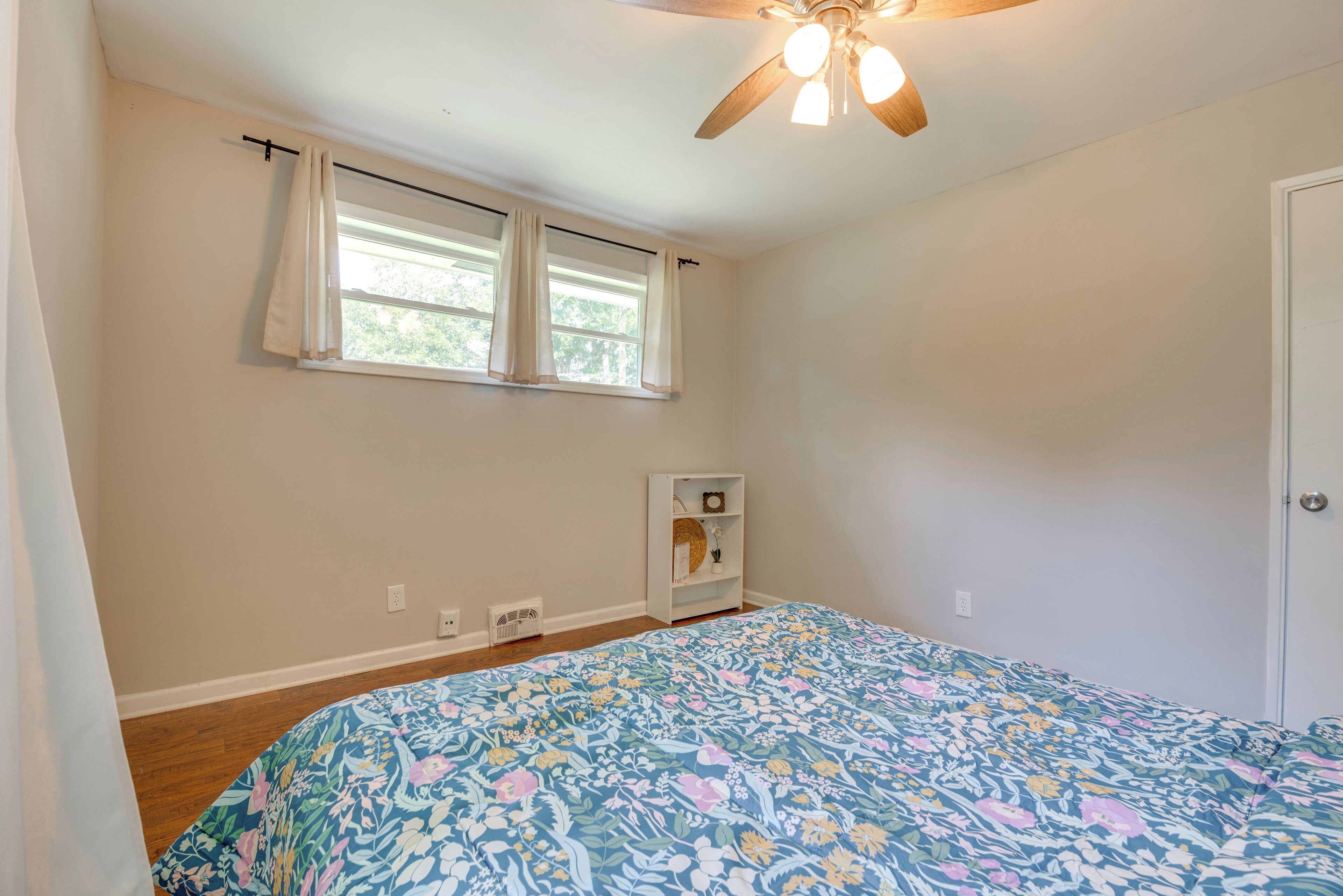 Quarto com cama, ventoinha no teto, janela com cortinas, prateleira branca e porta.