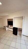 Quarto com escrivaninha branca, geladeira preta e televisão na parede.