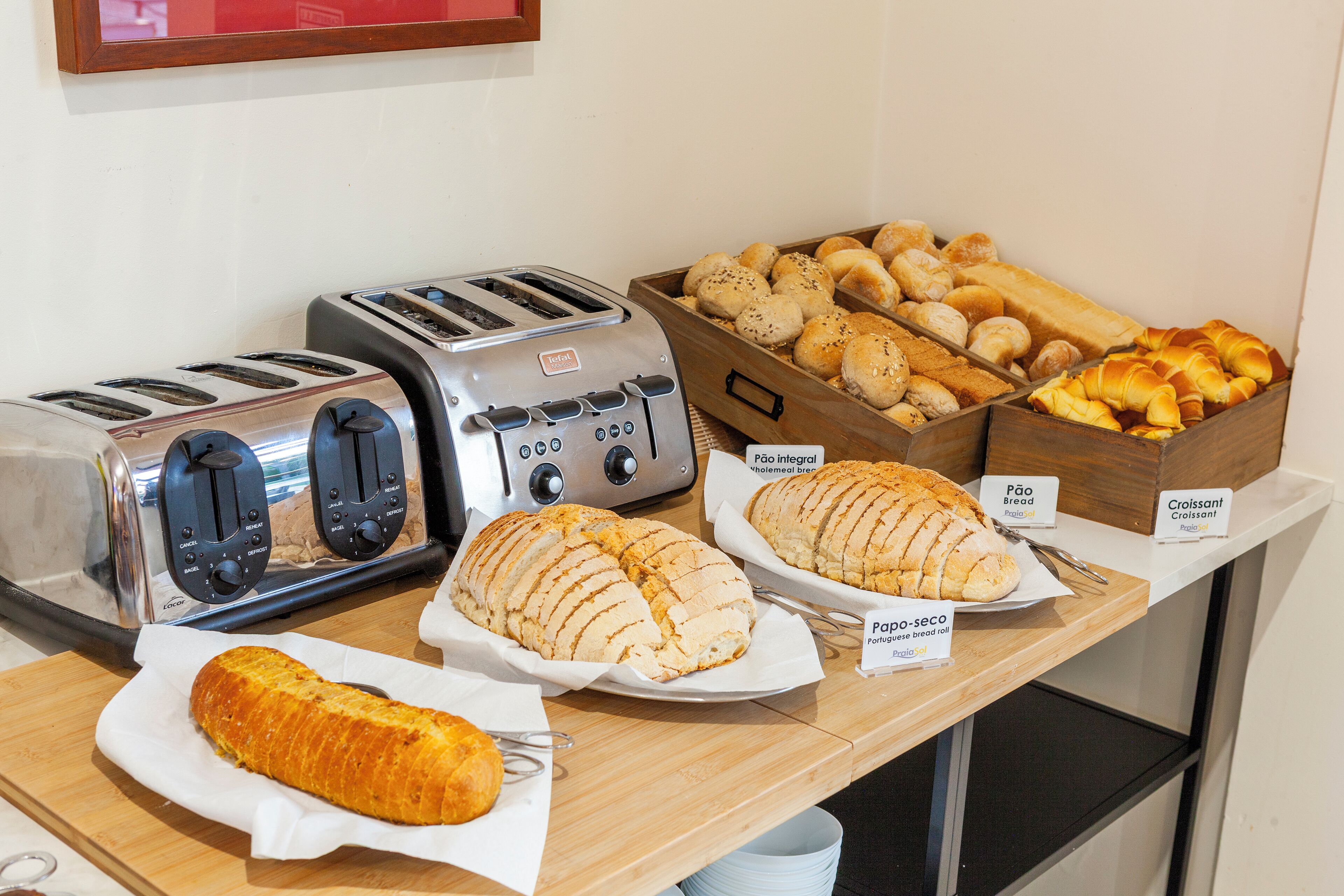 Buffet de café da manhã com diversos pães e doces, incluindo pão integral, pão francês, croissant e pão de forma seco.