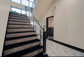 Uma escadaria com degraus pretos e corrimão de metal, que leva a um quarto com parede branca e uma porta.