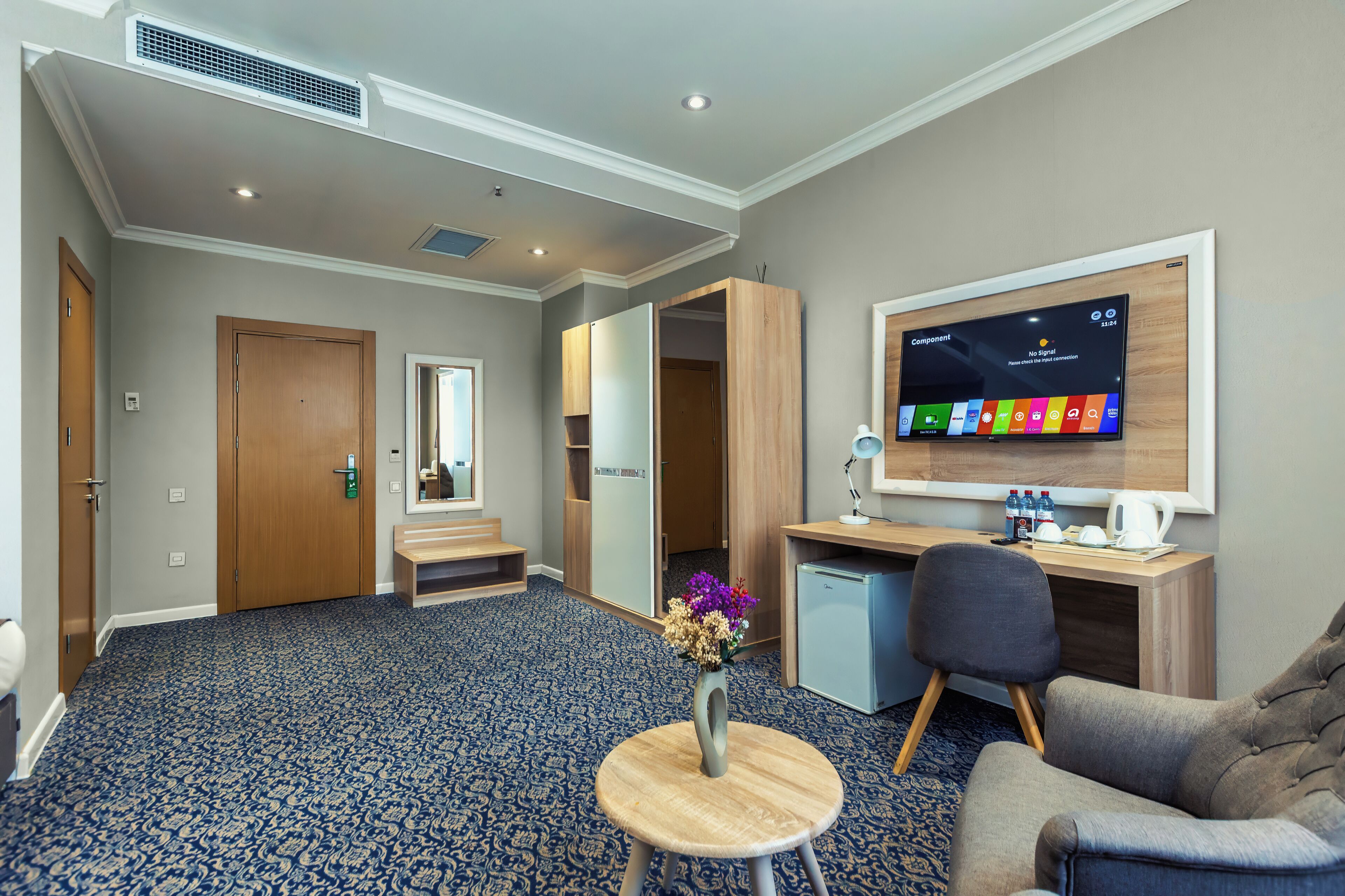 Quarto de hotel com uma televisão de tela plana grande fixada na parede, uma escrivaninha com cadeira, um pequeno refrigerador, um banco e um tapete estampado.