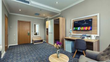 Quarto de hotel com uma televisão de tela plana grande fixada na parede, uma escrivaninha com cadeira, um pequeno refrigerador, um banco e um tapete estampado.