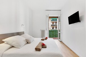 Quarto de hotel moderno com uma cama bem arrumada, televisão de tela plana na parede e vista para um edifício com varandas através de uma porta aberta.