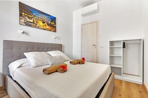 Cama bem arrumada com lençóis brancos e duas toalhas enroladas com flores vermelhas, um quadro com vista urbana acima, e um guarda-roupa com prateleiras abertas.