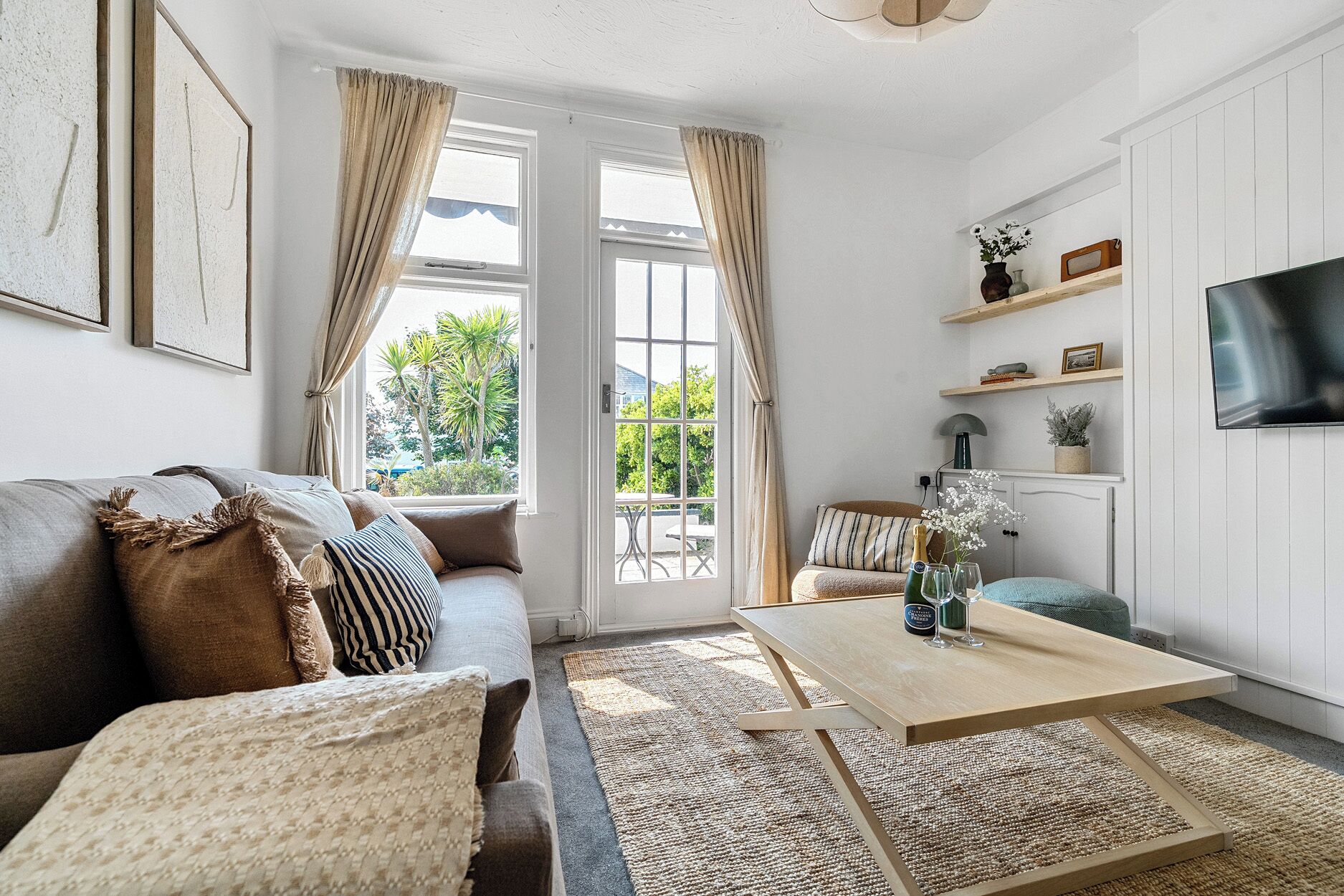 Uma sala de estar aconchegante com sofá, mesa de centro e TV na parede. Há cortinas nas janelas e uma pequena estante com objetos decorativos.