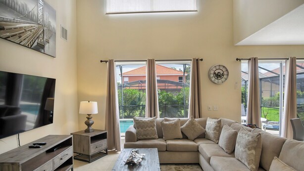 Uma sala de estar com uma grande televisão de tela plana, uma mesa de centro de madeira, um sofá bege com almofadas estampadas e vista para uma piscina e uma casa através de grandes janelas.