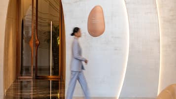 Entrada de um edifício moderno com porta de vidro, parede curva e placa indicando o hall dos elevadores.