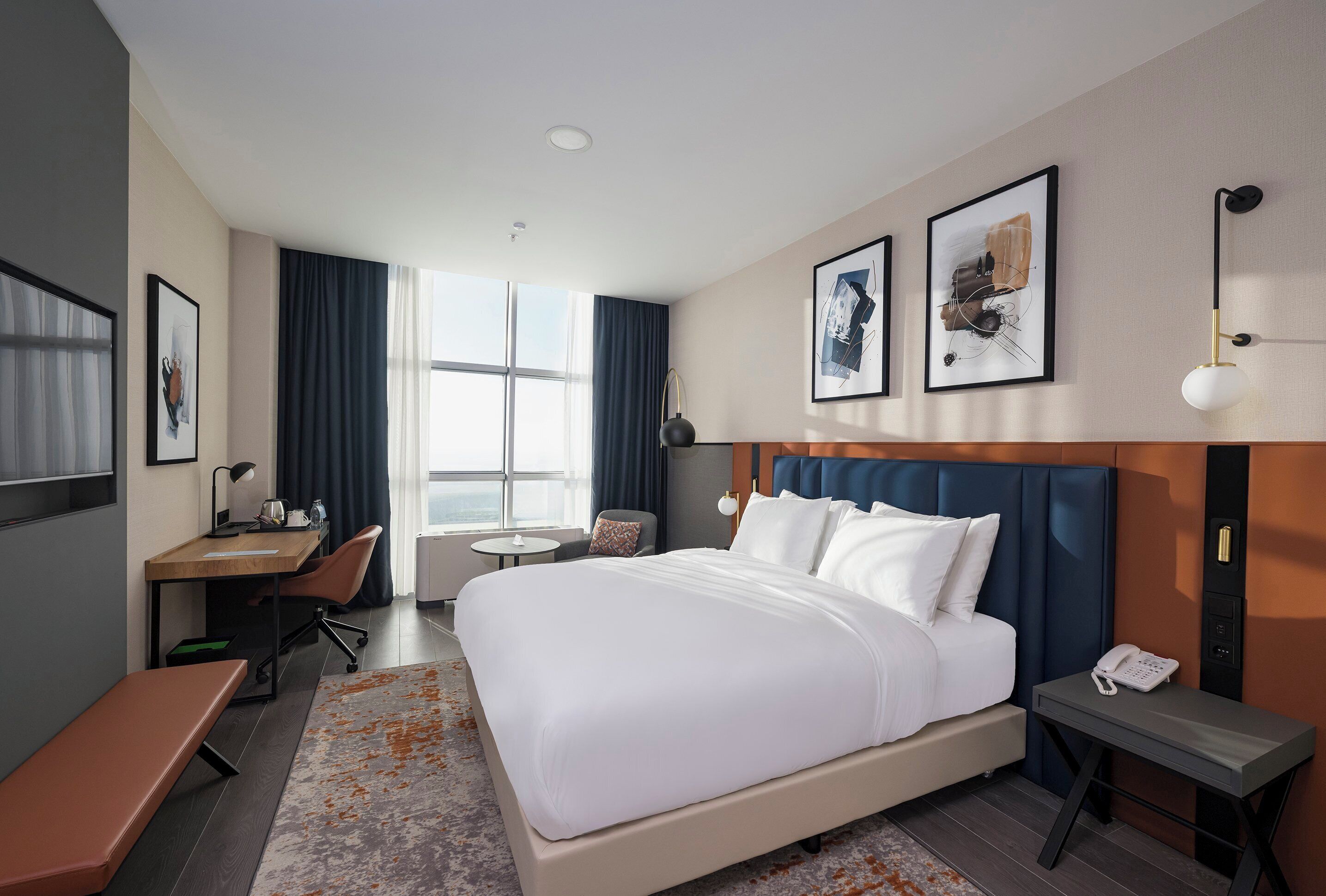 Quarto de hotel com cama, escrivaninha, cadeira, mesa pequena e telefone num criado-mudo.