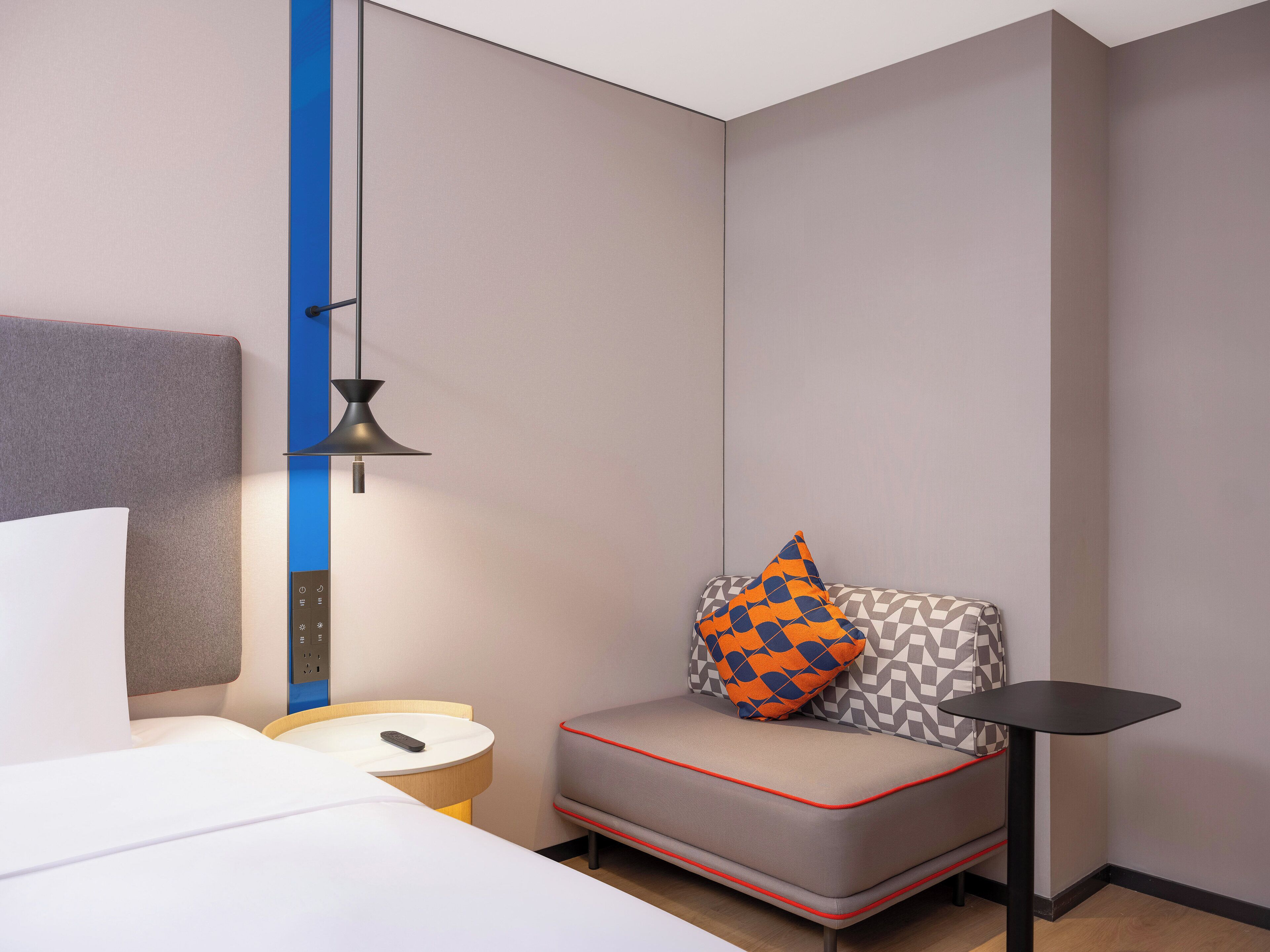 Quarto de hotel moderno com cama, mesa pequena, sofá com almofadas estampadas e luminária pendente.