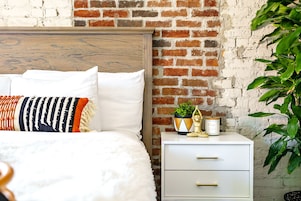 Uma cama com roupa de cama branca, um travesseiro listrado e uma mesa de cabeceira com uma planta e uma pequena estatueta.