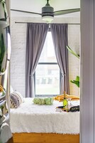 Quarto com cama macia e branca, ventilador de teto, janela com cortinas e vista para o exterior.