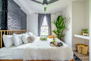 Quarto com cama, ventoinha no teto, janela com cortinas, planta e uma prateleira pequena com objetos decorativos.