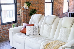 Um sofá de couro branco com encostos acolchoados e um cobertor, colocado em um quarto com paredes de tijolo e janelas grandes.