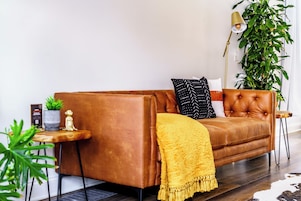 Um sofá de couro marrom com uma manta amarela, uma pequena mesa lateral com uma planta e um abajur alto.
