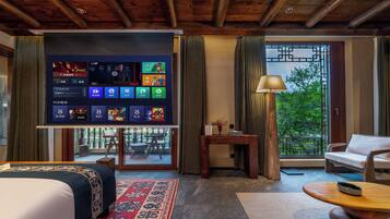 Quarto de hotel com uma televisão grande exibindo vários aplicativos, uma cama com colcha estampada, uma mesa de centro de madeira, um sofá e vista para o verde pela janela.