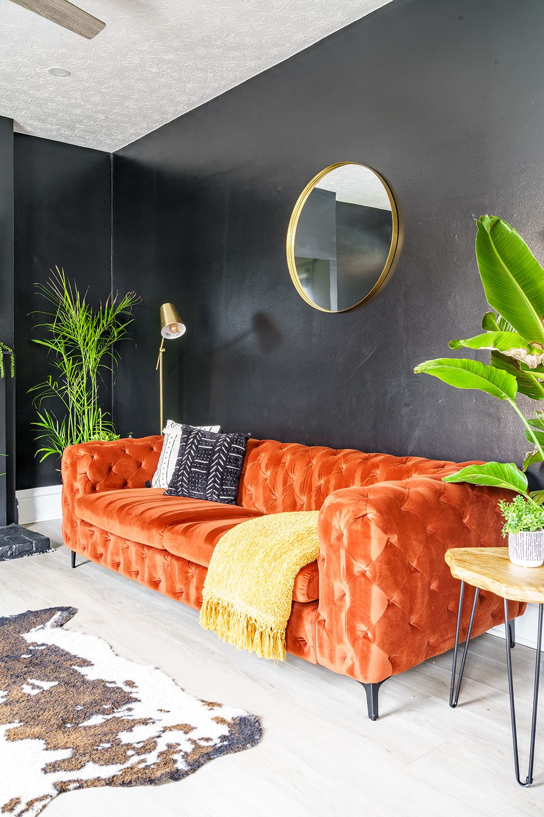 Uma sala de estar moderna com uma parede de destaque escura, um sofá laranja com capitonê, um espelho redondo e uma pequena mesa redonda com uma planta.