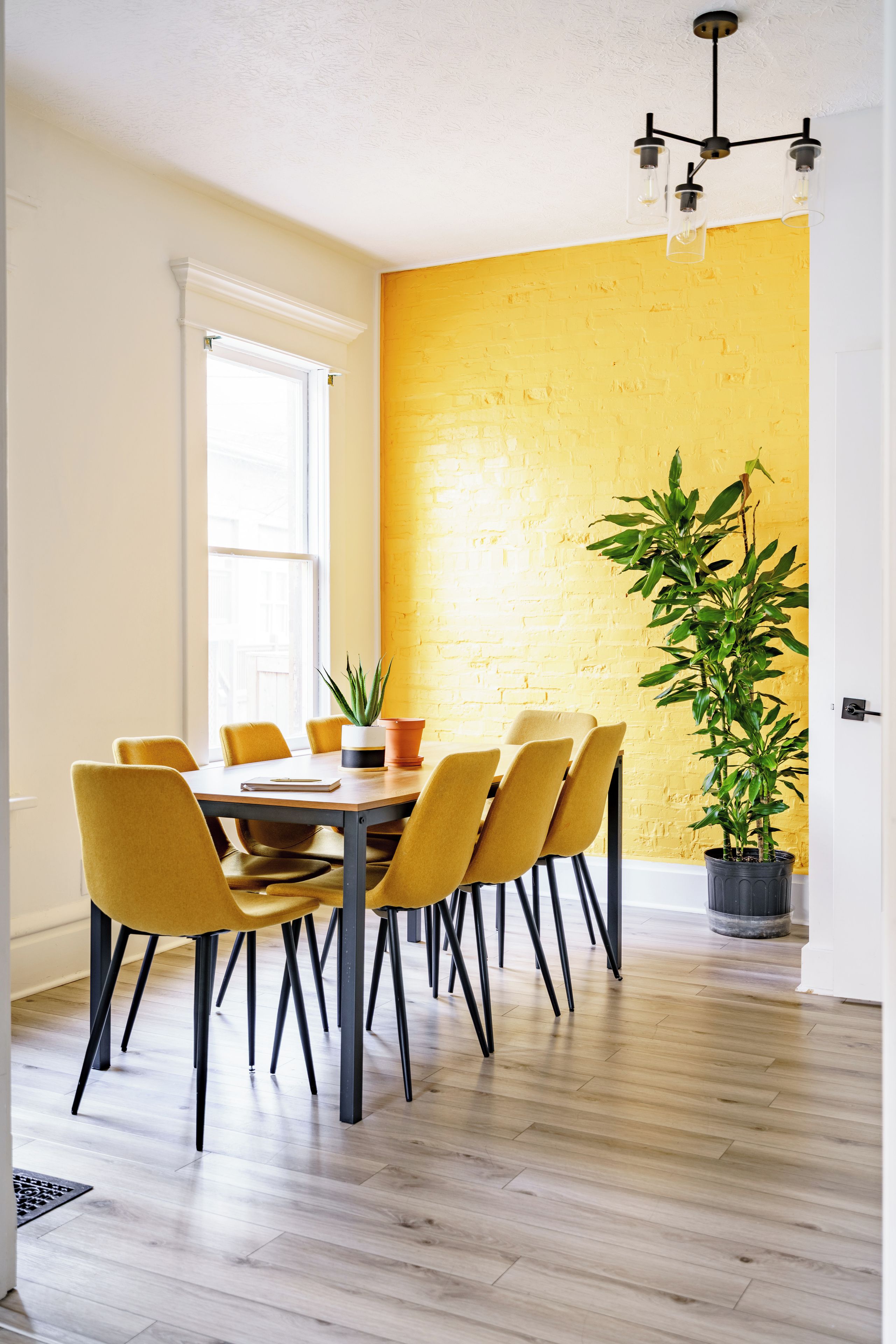 Uma área de refeições com mesa, cadeiras, uma planta em vaso e uma parede de tijolos amarelos.