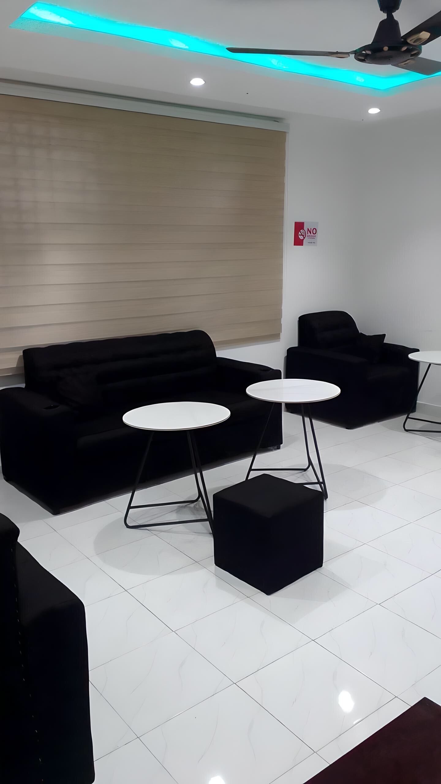 Uma área de estar moderna com sofás pretos e mesas redondas, um ventilador de teto e um aviso de 'Não Fumar'.