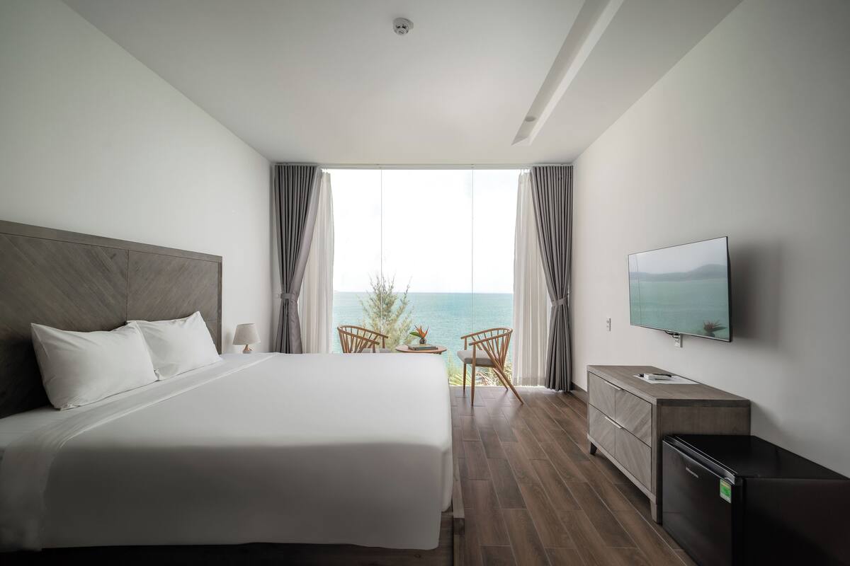 Quarto de hotel com uma cama grande, televisão, penteadeira de madeira e vista para o mar através de grandes janelas.