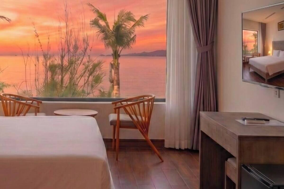 Quarto de hotel com cama, escrivaninha, cadeira e vista para o pôr do sol sobre a água.