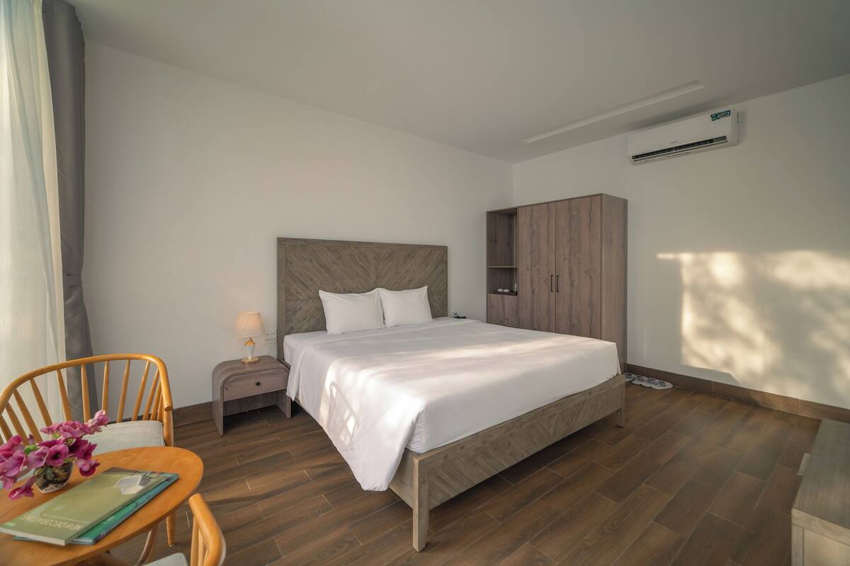 Quarto de hotel com cama de madeira, mesa de cabeceira, guarda-roupa de madeira, pequena mesa com abajur e uma cadeira.
