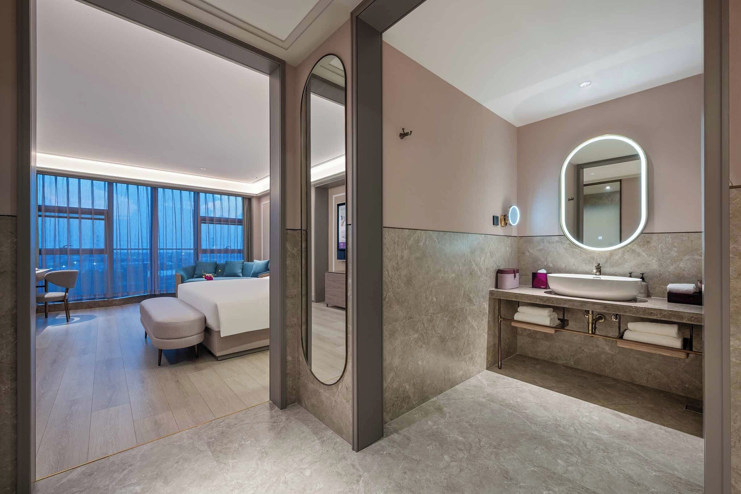 Quarto de hotel moderno com uma cama grande, área de estar, banheiro com toucador e espelho, e vista através de uma porta de vidro.