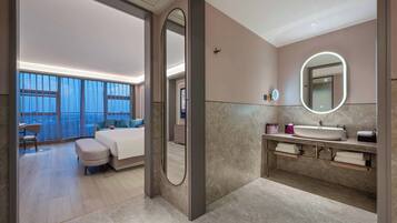 Quarto de hotel moderno com uma cama grande, área de estar, banheiro com toucador e espelho, e vista através de uma porta de vidro.