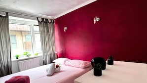 Quarto com cama, travesseiro rosa, toalha enrolada, pequena planta em vaso e um objeto preto no criado-mudo.