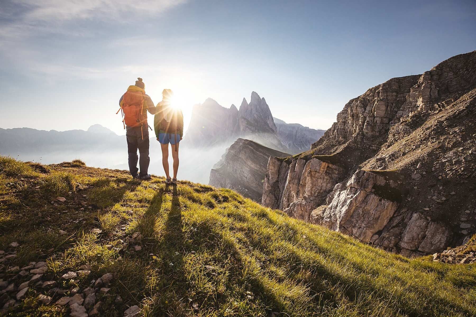 Duas pessoas caminhando por uma trilha gramada na montanha, com um penhasco rochoso de um lado e uma cadeia de montanhas ao fundo.