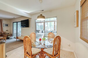 Uma área de refeições com mesa de vidro e cadeiras de vime, um sofá ao fundo e uma TV fixada na parede.