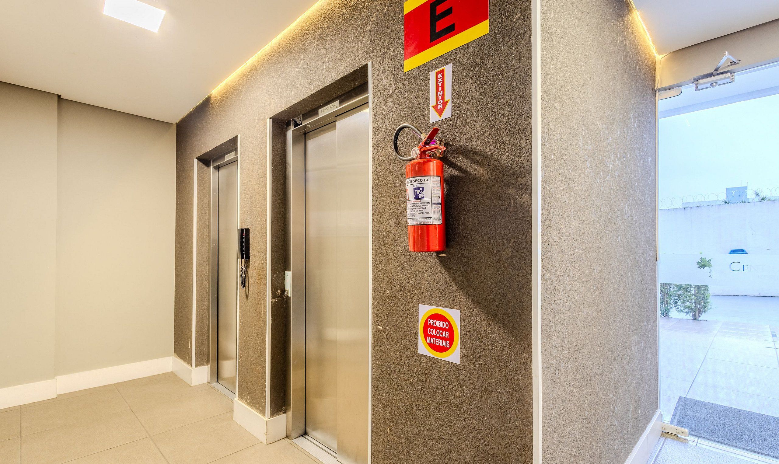 Corredor com dois elevadores, um extintor de incêndio e um aviso proibindo o depósito de materiais.