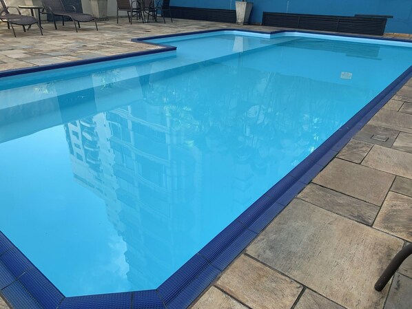 Piscina retangular com borda azul, cercada por ladrilhos de pedra e cadeiras de vime.