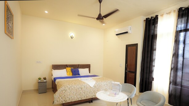 Quarto de hotel com cama, mesa de cabeceira, duas cadeiras, ventoinha no teto e janela com cortinas.