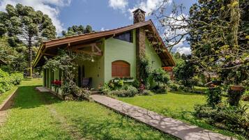 Uma casa com fachada verde, varanda de madeira e um jardim com diversas plantas e um caminho que leva à entrada.