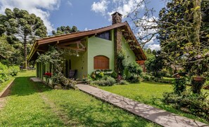 Uma casa com fachada verde, varanda de madeira e um jardim com diversas plantas e um caminho que leva à entrada.