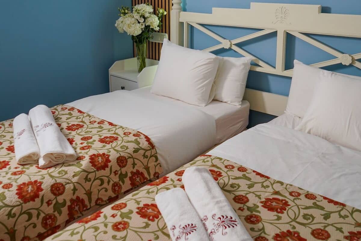 Quarto de hotel com duas camas, roupa de cama com padrão floral, almofadas brancas e parede azul.