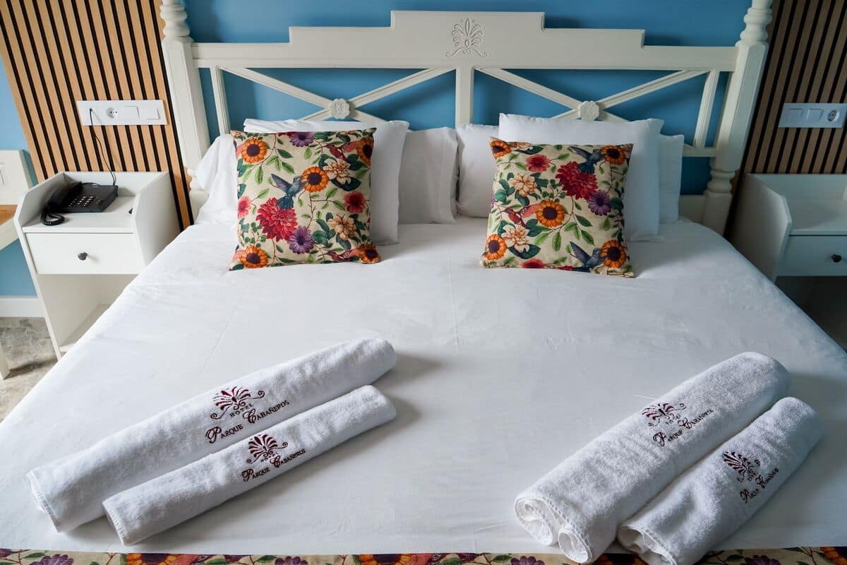 Uma cama bem arrumada com almofadas florais, uma mesa de cabeceira branca com um telefone e duas toalhas enroladas com bordado.