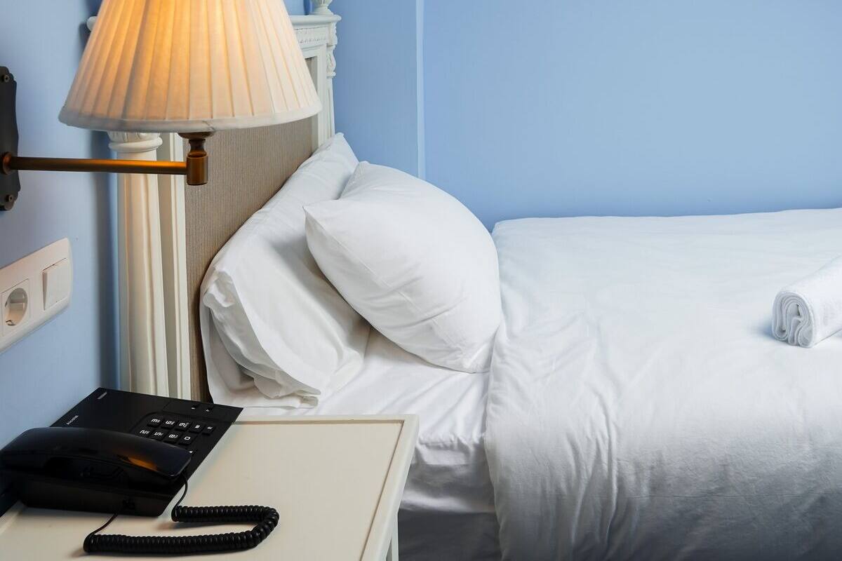 Quarto de hotel com cama, roupa de cama branca, abajur, telefone e tomada na parede.