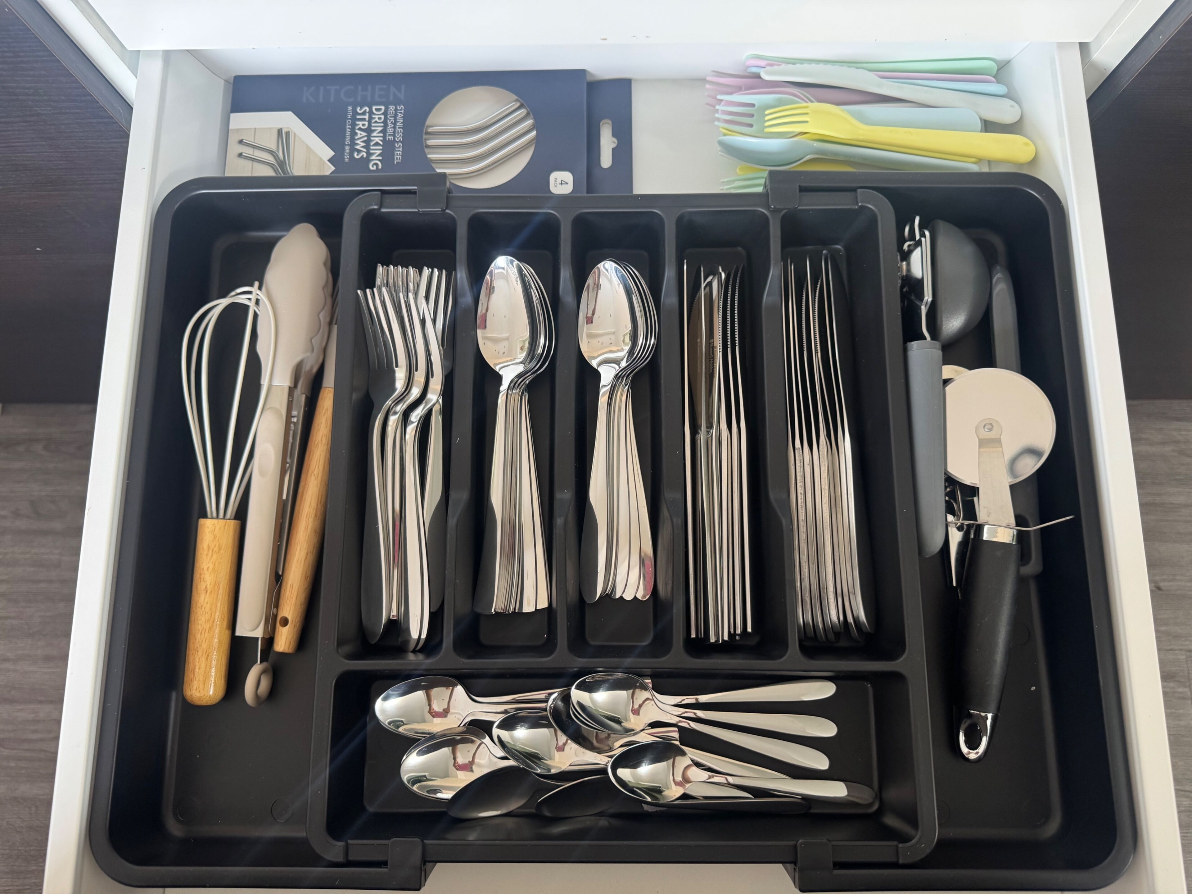 Uma gaveta de cozinha com utensílios organizados em compartimentos, incluindo batedores, garfos, colheres e um cortador de pizza.