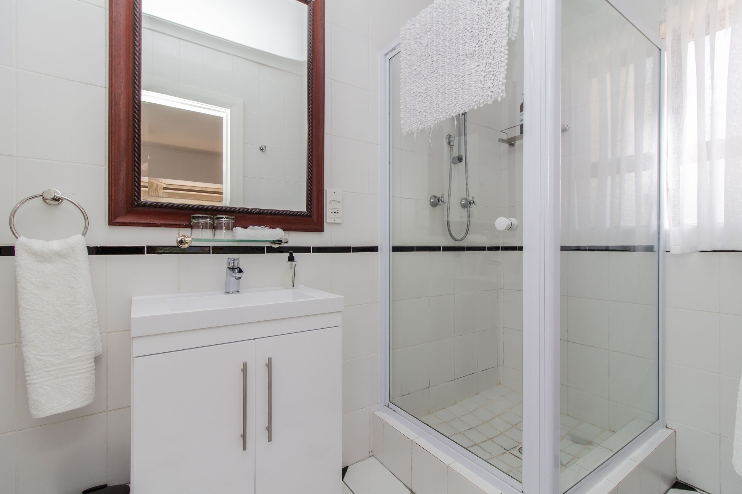 Casa de banho com chuveiro em cabine de vidro, lavatório branco com espelho em cima e toalha pendurada num suporte de metal.