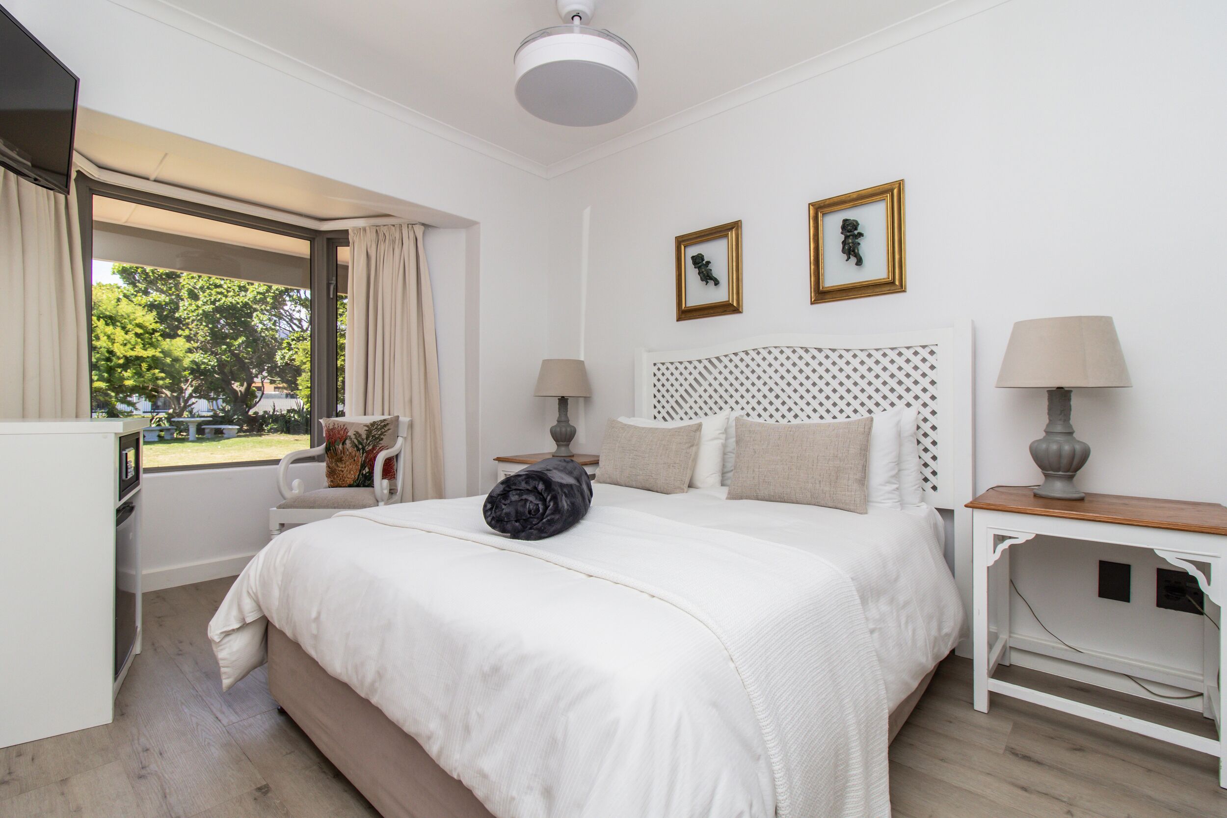 Um quarto bem arrumado com uma cama grande, duas mesas de cabeceira com luminárias, uma janela com cortinas e duas fotos emolduradas na parede.