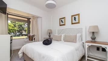 Um quarto bem arrumado com uma cama grande, duas mesas de cabeceira com luminárias, uma janela com cortinas e duas fotos emolduradas na parede.