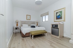 Quarto com cama, mesas de cabeceira, um banco, uma kitchenette pequena e uma janela.