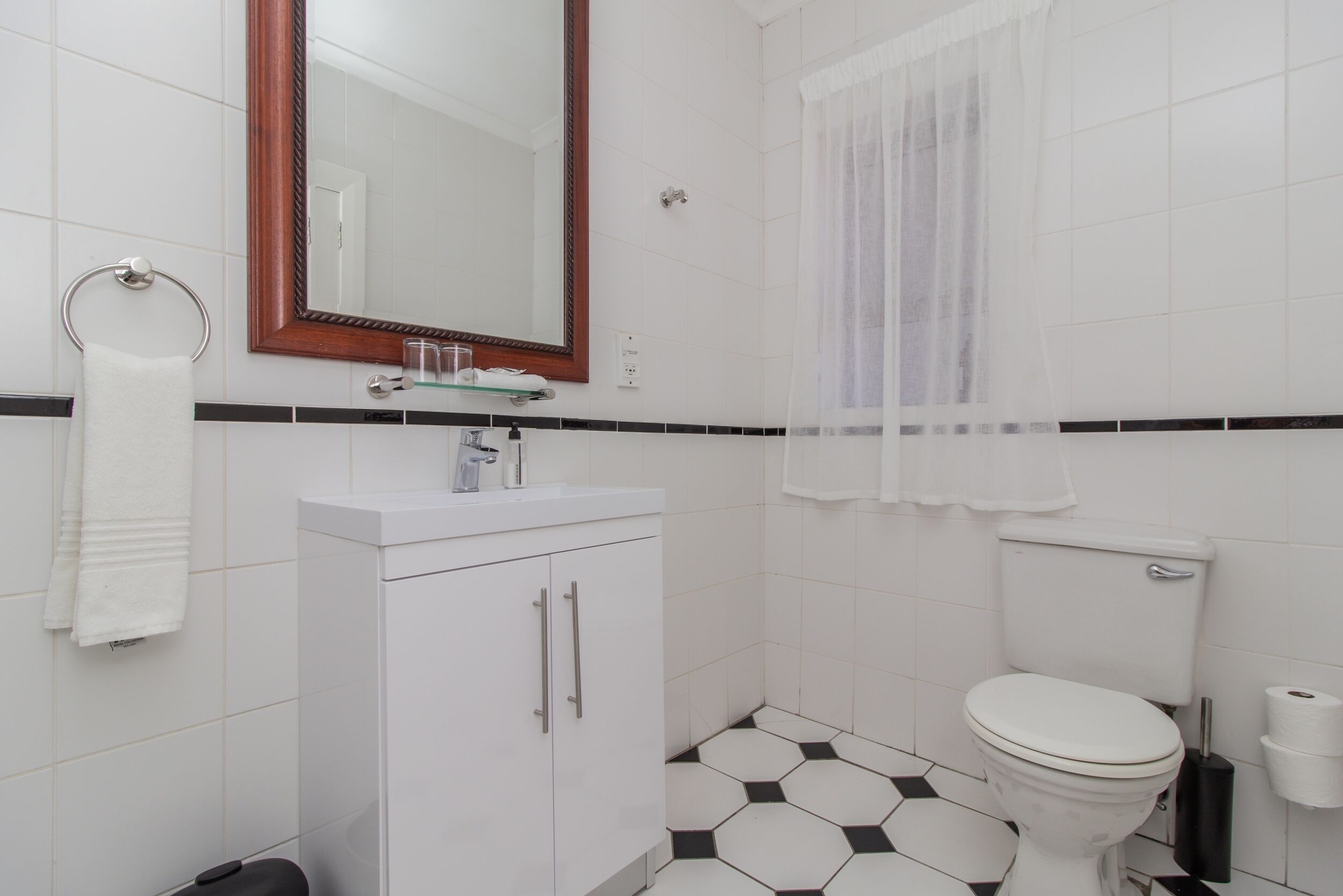 Casa de banho com lavatório branco, espelho, sanita e mosaicos hexagonais no chão.