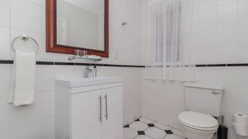 Casa de banho com lavatório branco, espelho, sanita e mosaicos hexagonais no chão.