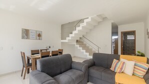 Uma sala de estar moderna com um sofá cinzento, uma área de refeições com cadeiras de madeira e uma mesa, e uma escadaria com guarda-corpo de vidro.