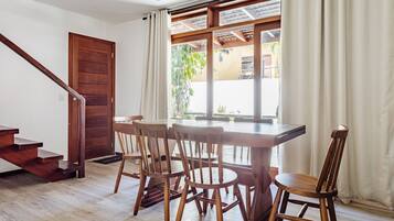 Uma área de refeições com mesa e cadeiras de madeira, uma escada de madeira e vista pela janela com cortinas.