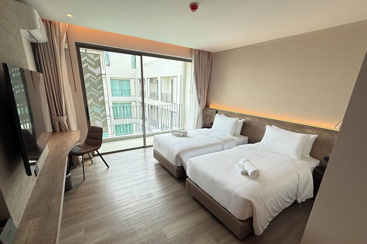 Quarto de hotel com duas camas, uma escrivaninha e uma cadeira. Há uma janela grande com vista para prédios.